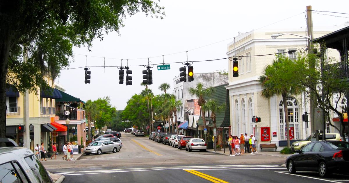 A bustling street corner showing pedestrians, cars, and a mix of classic and modern storefronts under a hazy sky.