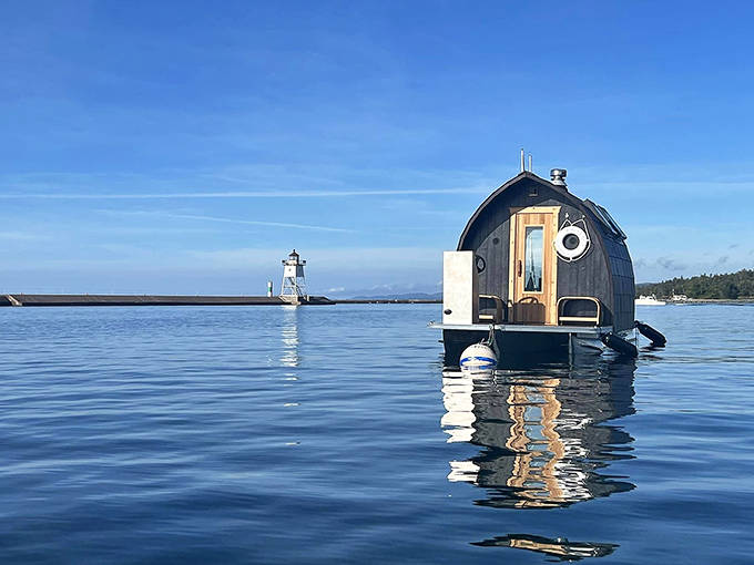 A wooden barrel-shaped sauna floats serenely on Lake Superior's crystal waters, with a lighthouse standing guard in the distance.