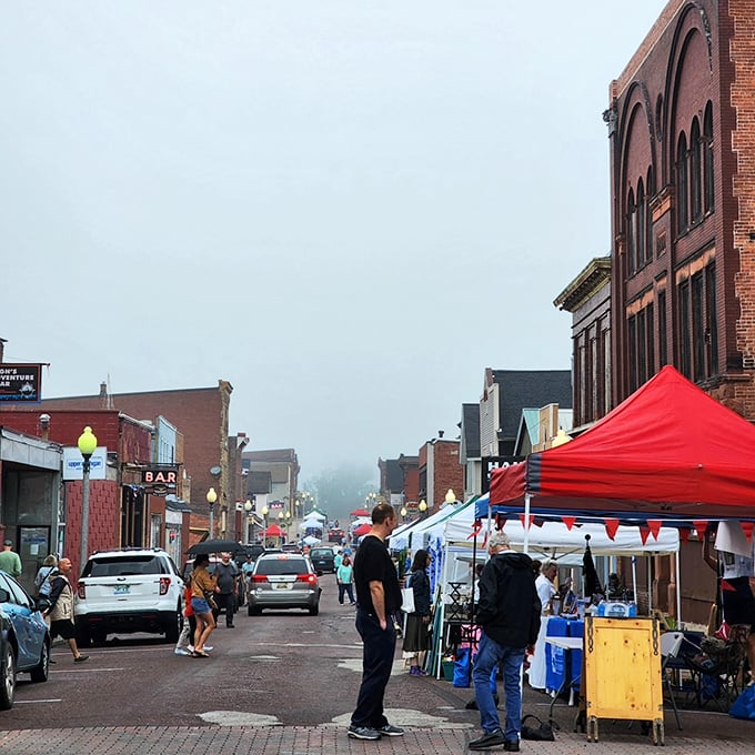 A cozy small-town street festival, with colorful tents lining brick buildings as people wander through the misty morning.