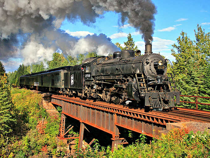 Steam billows dramatically as this vintage locomotive crosses the bridge, a magnificent iron horse against Minnesota's verdant backdrop.