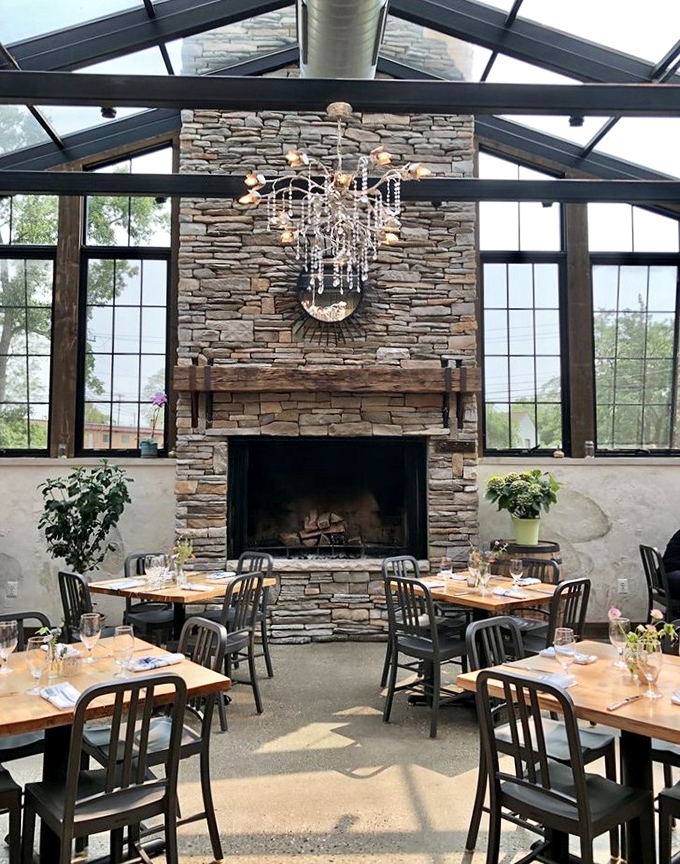 A stone fireplace anchors the sunlit dining room, promising cozy warmth during Michigan winters while tables await guests seeking exceptional farm-to-table cuisine.