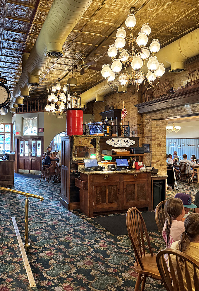 Elegant chandeliers illuminate the spectacular pressed tin ceiling while exposed brick walls create a warm embrace that feels like stepping into a sepia photograph.