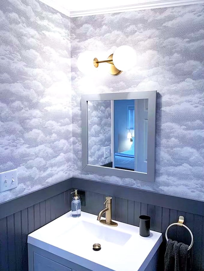 Cloud-patterned wallpaper and brass fixtures transform this bathroom into a dreamy maritime sanctuary worthy of Neptune himself.