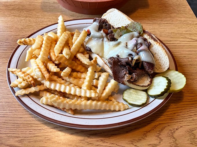 A hearty Philly cheesesteak loaded with sliced beef, melted cheese, and peppers, paired with crinkle-cut fries and dill pickles.
