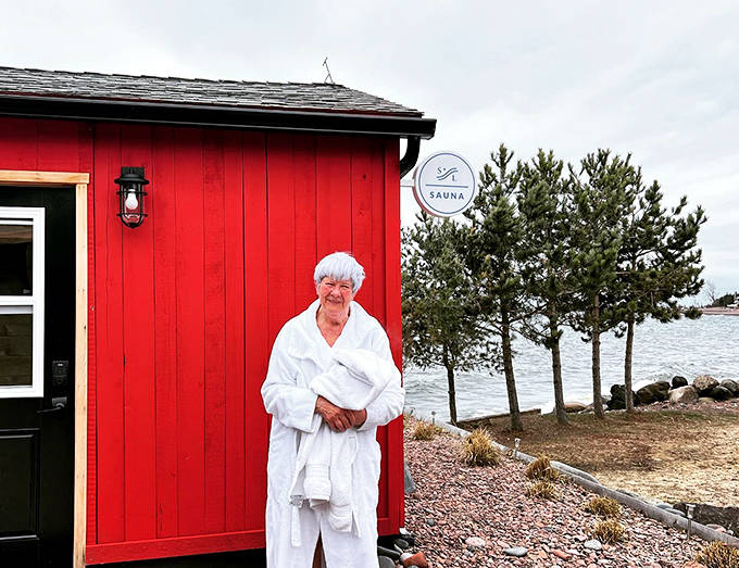 A person wrapped in white stands beside the bright red sauna building, looking like they've discovered the secret to Minnesota winter survival.