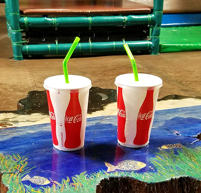 Two Coca-Cola cups with bright green straws sit like old friends – simple pleasures that taste inexplicably better after working up a playground sweat.
