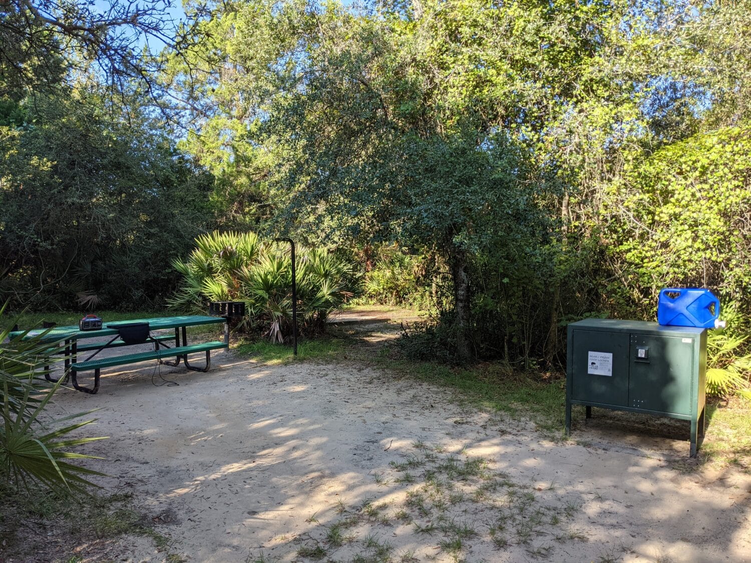 a campsite setup with a picnic table grill and a portable water container surrounded by lush vegetation