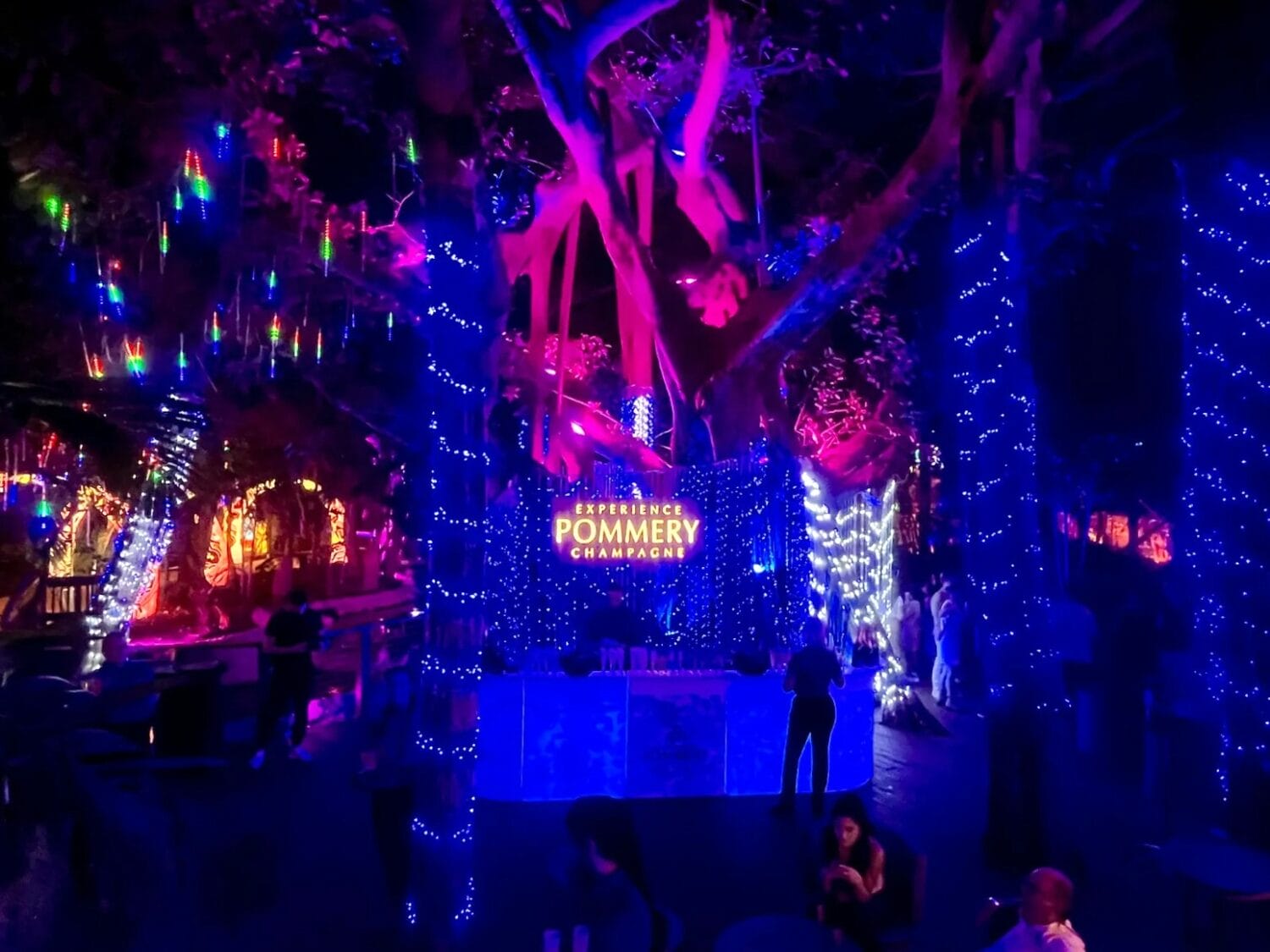 a festive outdoor setting with trees wrapped in blue lights and hanging colorful raindrop shaped decorations creating an enchanting evening atmosphere