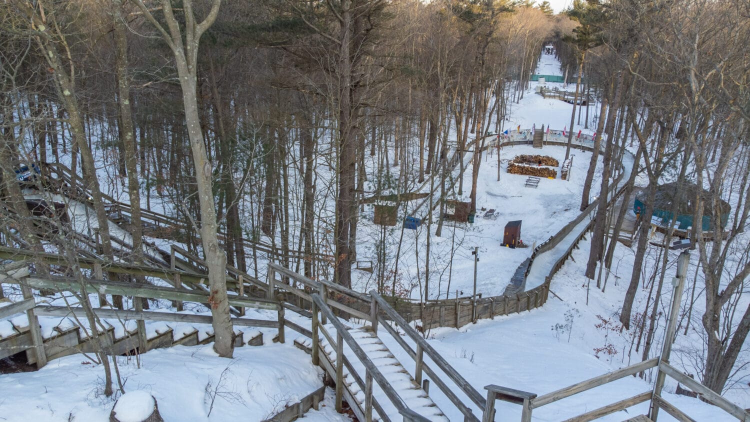 a tranquil view of a luge track in the winter showing the track winding through a forested area with snow covered trees and a stack of firewood in the foreground