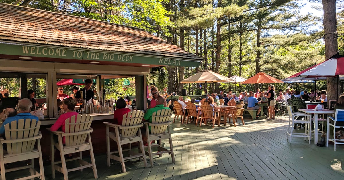 a bustling outdoor deck at Boone&rsquo;s Long Lake Inn with guests enjoying meals and drinks under the shade of umbrellas, surrounded by tall pine trees.