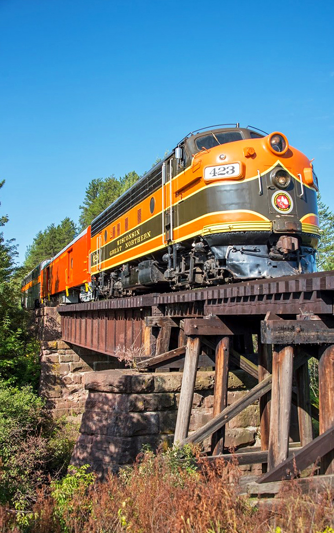 The majestic orange and yellow Wisconsin Great Northern locomotive powers across a wooden trestle, a splash of vintage color against the green wilderness.