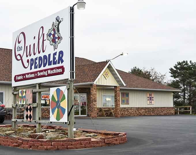 The welcoming storefront of The Quilt Peddler stands like a beacon for fabric enthusiasts, colorful quilt blocks hinting at treasures inside.