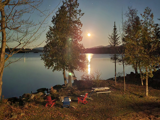 Moonlight creates a silver pathway across still waters, while Adirondack chairs stand ready for stargazers seeking cosmic perspective and nighttime conversations.