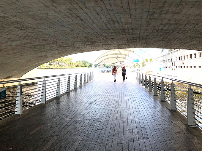 Sunlight filters through the bridge, creating dramatic shadows on the Riverwalk's path as pedestrians enjoy this architectural feature.