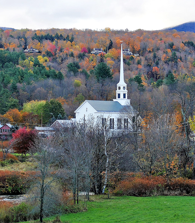 A classic white steeple rises above autumn's fiery display, embodying the quintessential New England scene that makes Vermont postcards famous worldwide.