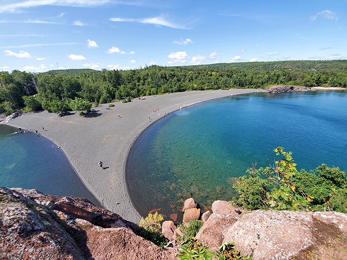 From above, the beach's crescent shape and dark sand create a striking contrast with the surrounding forest and brilliant blue water.