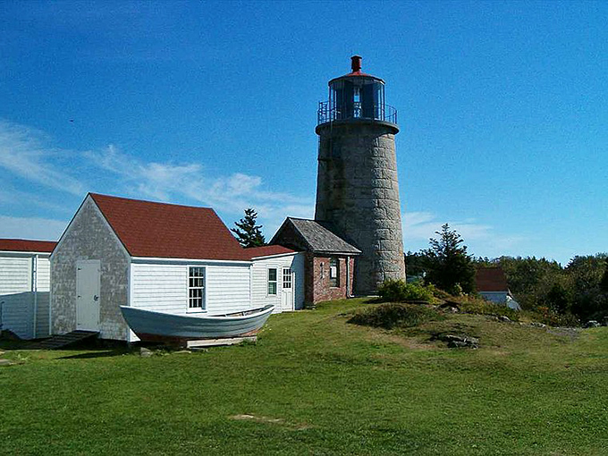 Monhegan's historic lighthouse stands tall against the blue sky, with white buildings and a wooden boat completing this quintessential Maine coastal scene.