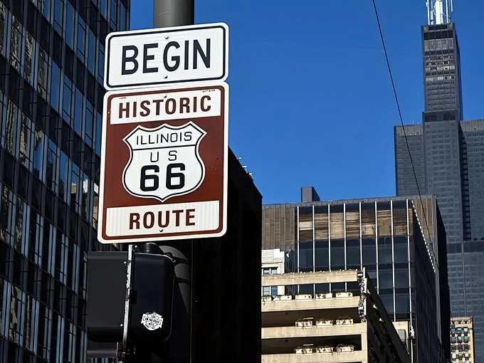 Skyscrapers tower over this humble brown shield sign where millions of Route 66 adventures have begun since 1926.