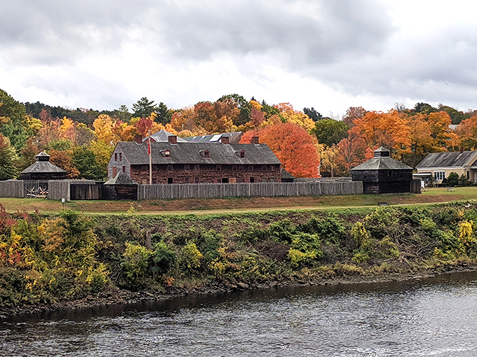Fall foliage creates a stunning backdrop for Old Fort Western, where the rustic buildings seem to glow against autumn's fiery palette along the riverbank.