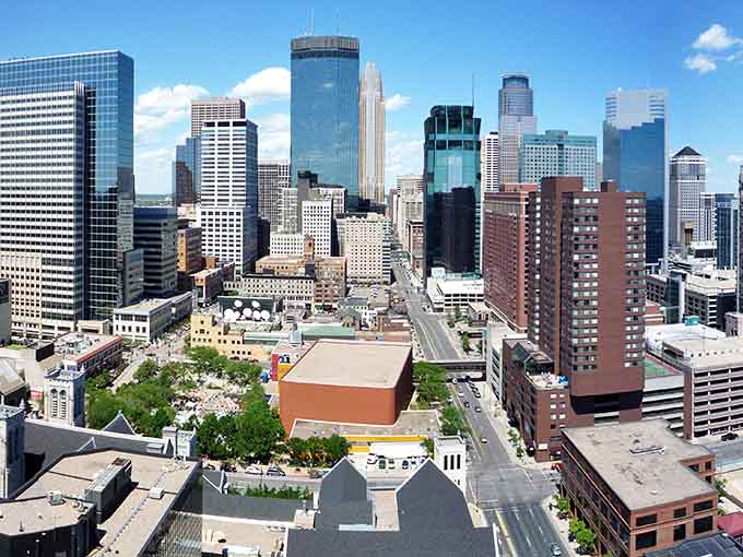 Minneapolis rises confidently into the sky, its modern buildings reflecting clouds and sunshine while the river flows peacefully below the bustling streets.
