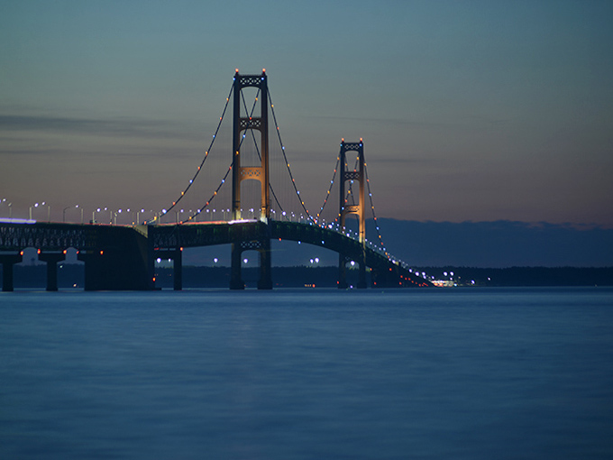 As twilight descends, the Mighty Mac transforms into a glittering necklace of lights, its illuminated span creating a magical pathway between worlds.