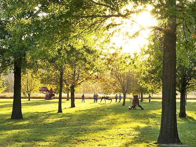 Golden hour bathes the sculpture garden in warm light, figures silhouetted against the trees like dancers frozen mid-performance.