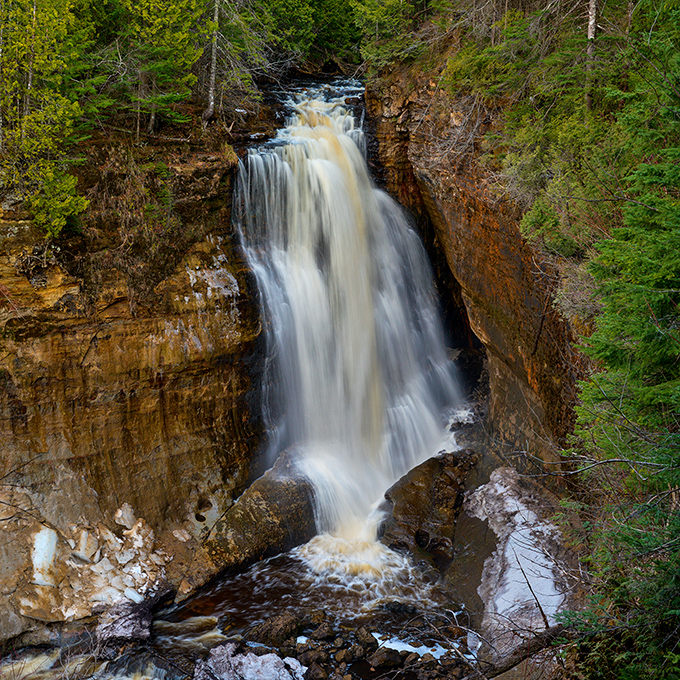 Miners Falls plunges dramatically between rocky walls, creating a misty curtain that catches sunlight filtering through the surrounding pine forest.