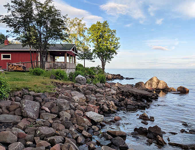 A charming red cabin sits among the rocks at Beaver Bay, where towering pines meet Lake Superior's crystal waters in perfect North Shore harmony.