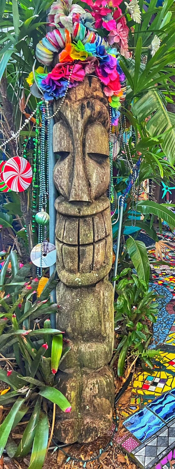 This wooden sentinel wears its floral crown with stoic dignity, Mardi Gras beads cascading down like a tribal elder's ceremonial garb.