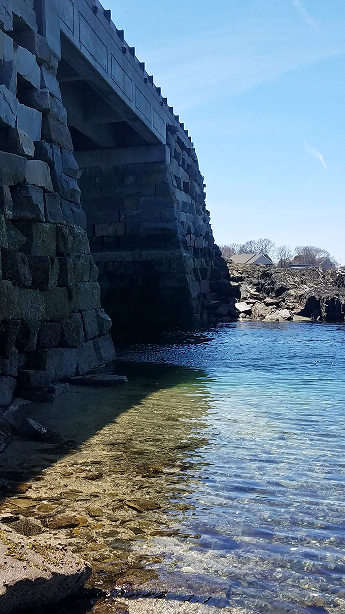 Dramatic perspective of the bridge's stonework against crystal-clear Maine waters, showcasing engineering that works with nature.