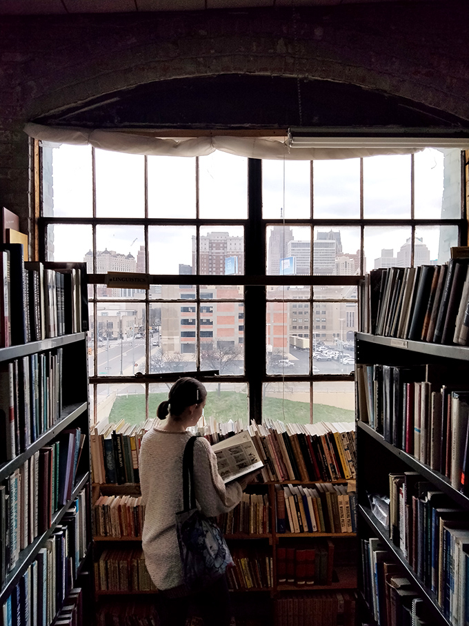 Standing before industrial windows framing the city beyond, this visitor experiences the perfect blend of solitary contemplation and urban connection through literature.