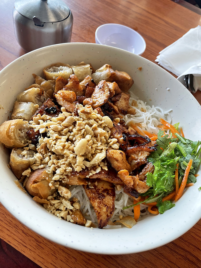 The Bun Ga Nuong Cha Gio combines grilled chicken, crispy egg rolls, and vermicelli noodles topped with crushed peanuts for textural paradise.