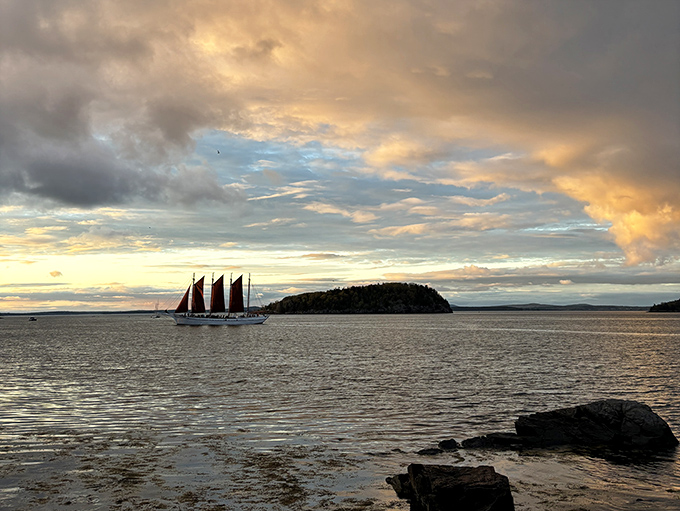 A crimson-sailed schooner glides across golden waters as the day's final light paints the sky in impossible colors.