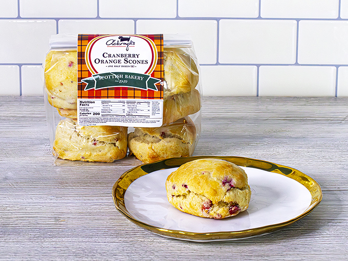 Fresh cranberry orange scone on a plate, with packaged bakery scones behind it, clean kitchen backdrop, bright and inviting display.