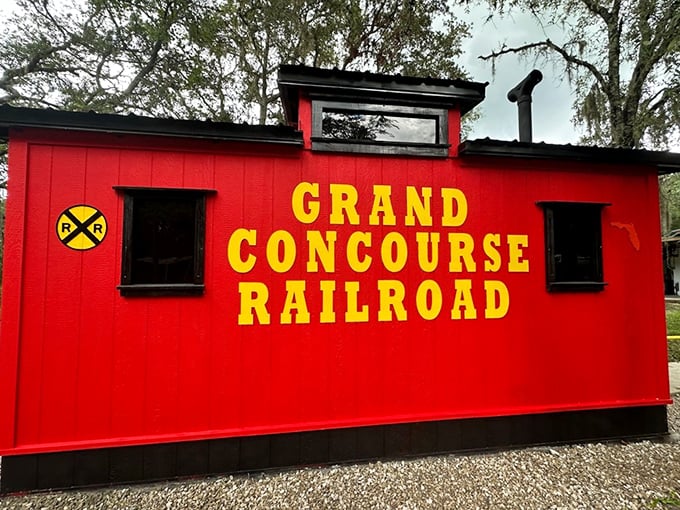 The vibrant red caboose of Grand Concourse Railroad stands proudly against Florida's greenery, its yellow lettering promising adventures bigger than its size suggests.