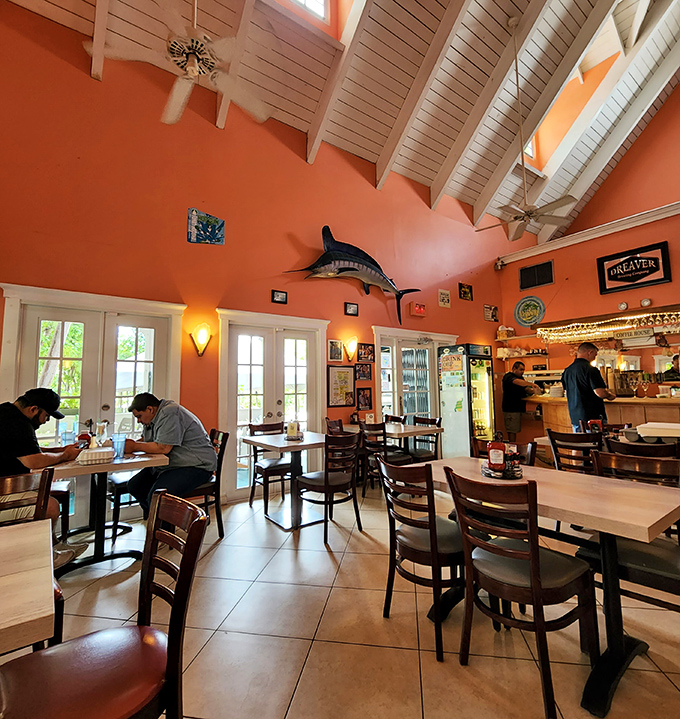 High coral walls and white ceiling beams create a space where conversations flow as easily as the ceiling fans spinning gentle reminders to relax.