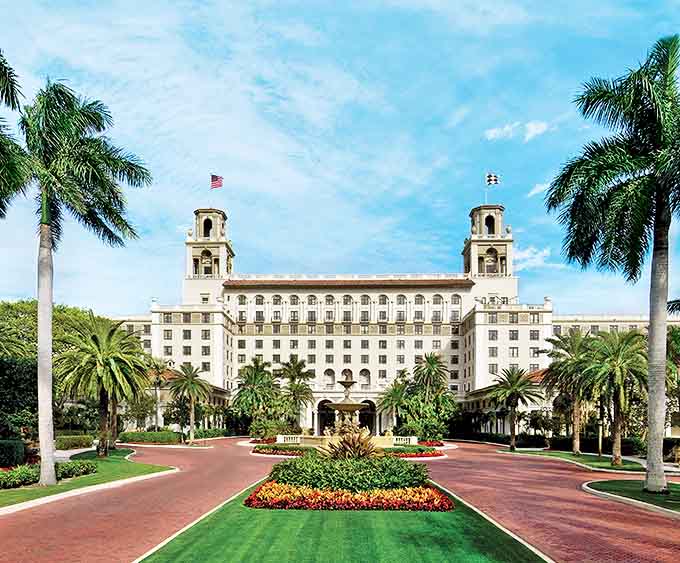 The Breakers' grand white facade stands majestically against blue skies, palm trees lining the entrance to this Palm Beach icon.