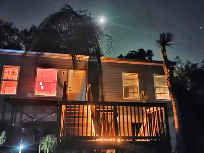 As darkness falls, the stilt house transforms into a glowing lantern, with warm light spilling from windows onto the moonlit landscape below.