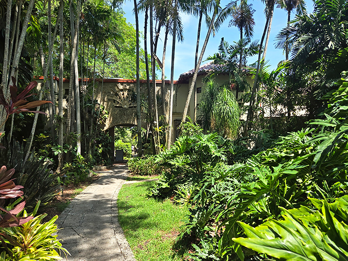 A verdant pathway leads through The Kampong's lush tropical landscape, where palm fronds create natural archways dappled with golden sunlight.