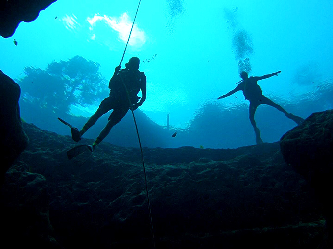 Divers explore the mysterious blue depths, silhouetted against sunlight streaming through the spring's underwater entrance.