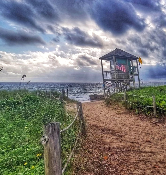 A sandy path leads past the lifeguard tower toward the shoreline, framed by sea grass and dramatic skies.