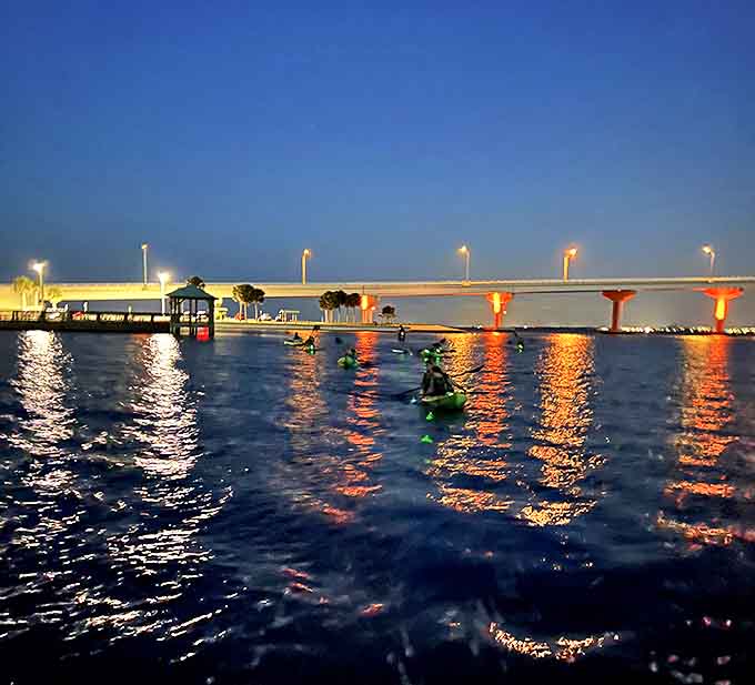 The bridge lights create a golden backdrop as kayakers with glowing safety gear prepare to explore the dark waters beyond.