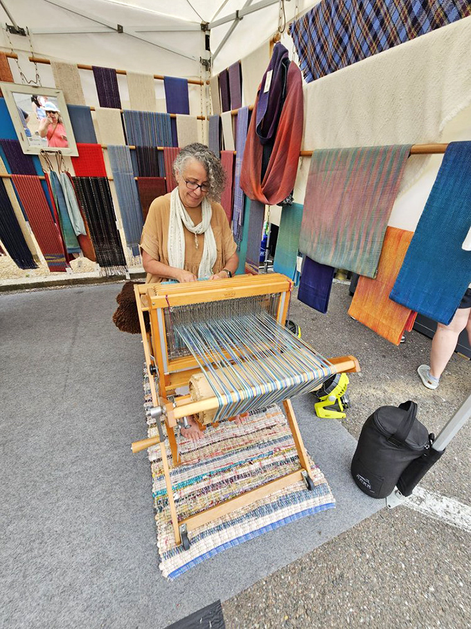 A skilled weaver demonstrates her craft amid a rainbow of handwoven textiles, her loom creating tomorrow's heirlooms before an appreciative audience.