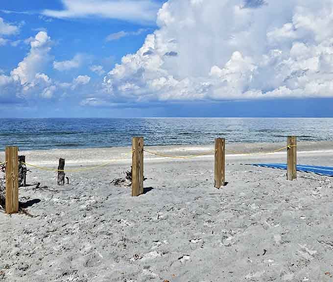 Rustic wooden posts and rope lines border the wide sandy beach under a dramatic sky filled with soft white clouds.