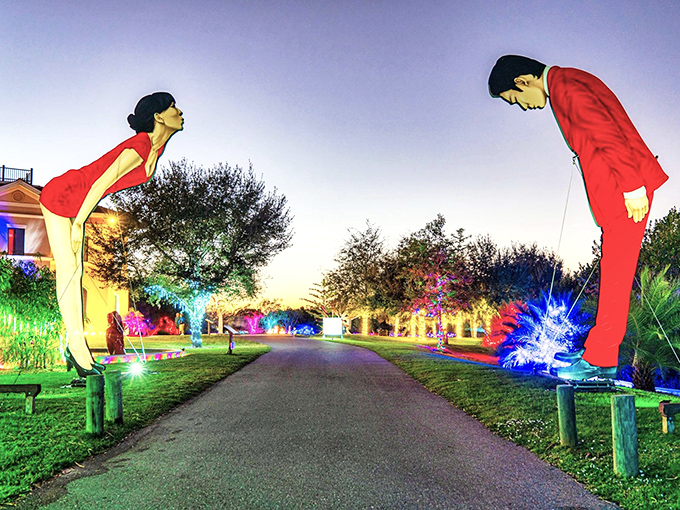 Giant sculptures bow in greeting at the garden entrance, illuminated by colorful lights creating a magical evening pathway.