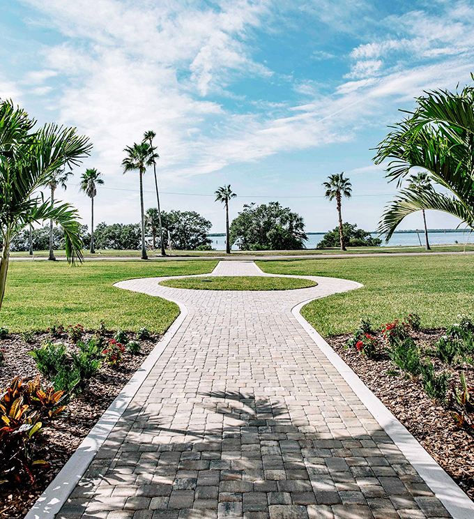 A brick pathway leads through manicured grounds toward the water, palm trees standing like exclamation points against the sky.