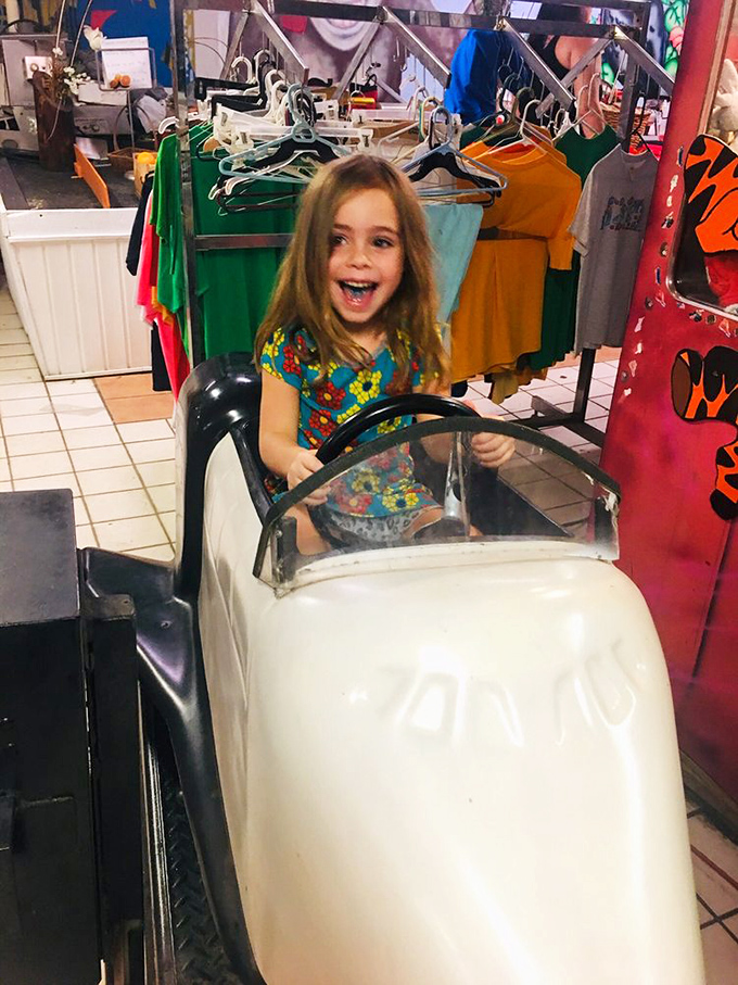Pure joy radiates from this young driver at the wheel of a vintage kiddie car, surrounded by the colorful chaos of the gift shop.
