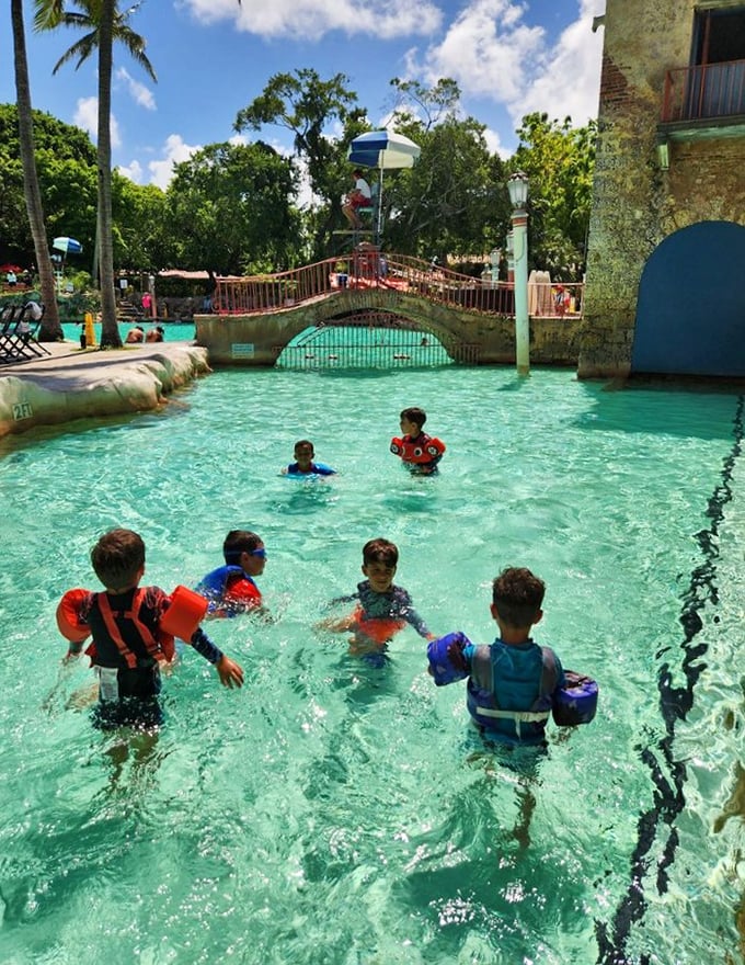 Young swimmers in bright life vests navigate the shallow areas, their laughter echoing off coral walls that have witnessed nearly a century of summer joy.