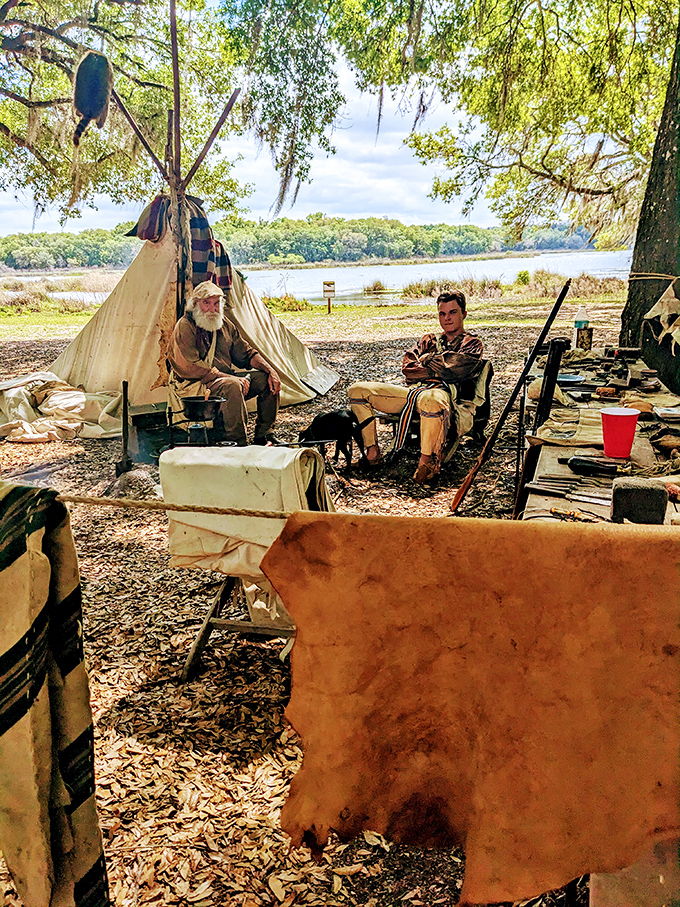Two period-dressed reenactors share tales by their authentic camp, the lake shimmering behind them like a liquid timeline.