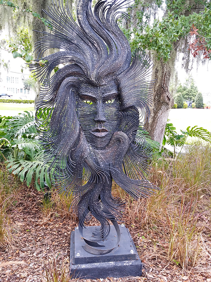 This dramatic metal face sculpture with flowing hair emerges from the landscape, its piercing eyes seeming to follow visitors through the garden.